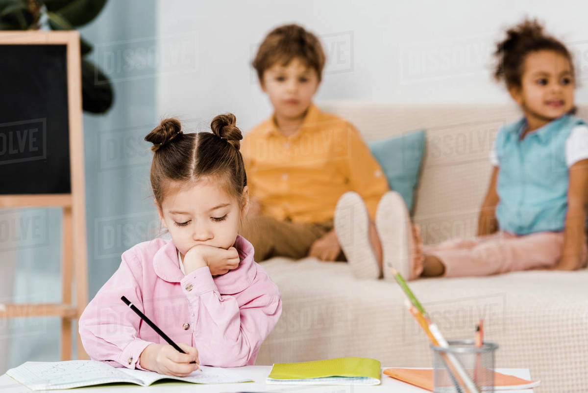 Adorable little child writing with pencil while friends sitting behind ...
