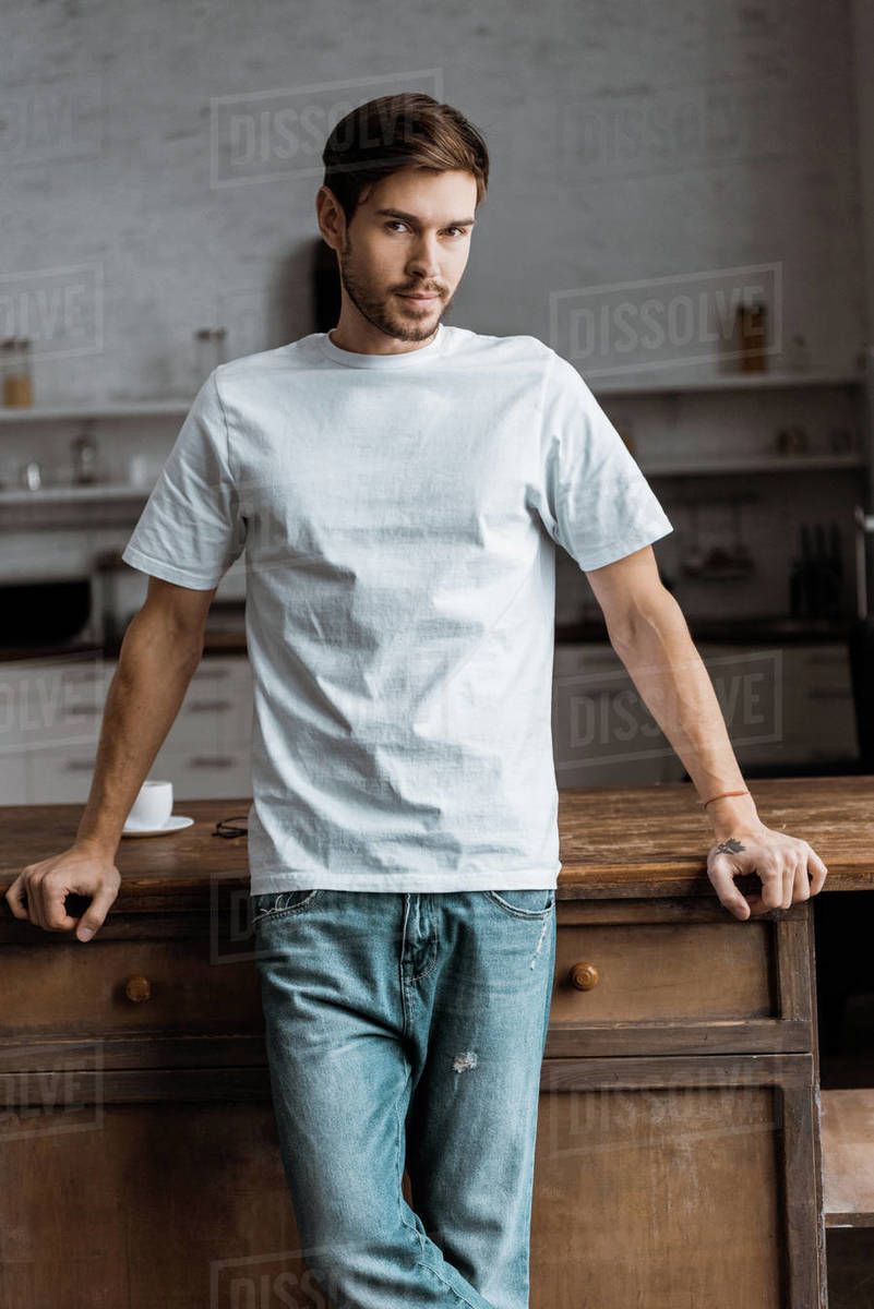 handsome young man leaning back on kitchen table and looking at camera ...
