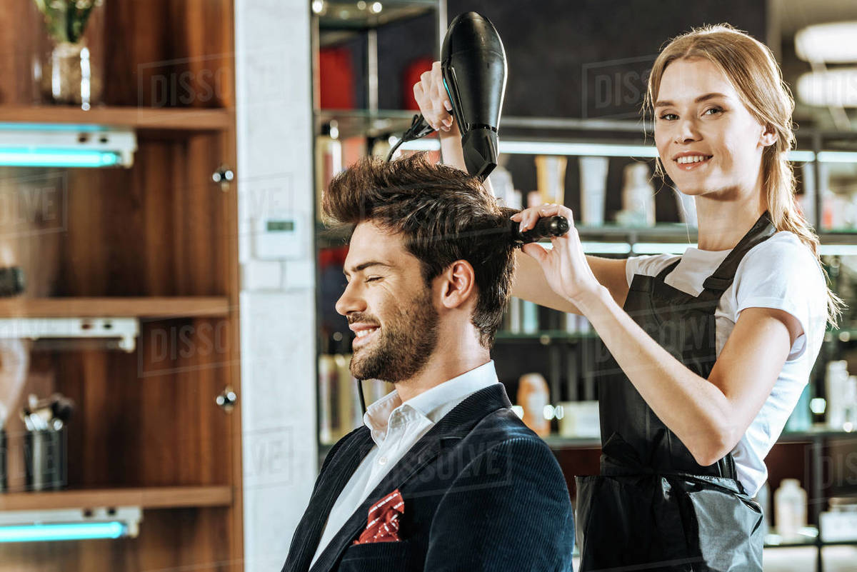 beautiful young hairstylist smiling at camera while drying and combing ...