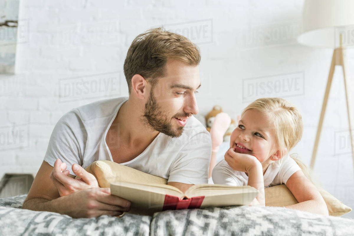 Happy Father And Daughter Smiling Each Other While Reading Book On Bed