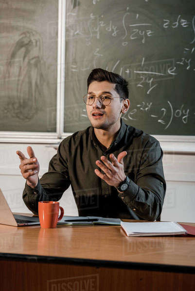 Male teacher in glasses sitting at desk and explaining equations in ...