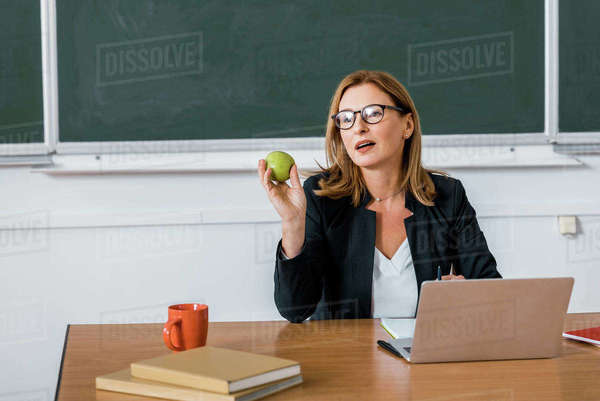 Female teacher sitting at computer desk and holding apple in classroom ...