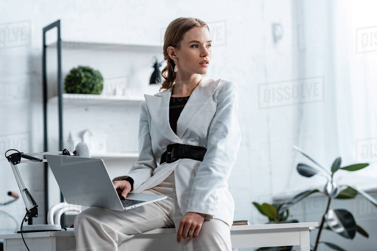 Stylish businesswoman in white formal wear sitting on desk with laptop ...