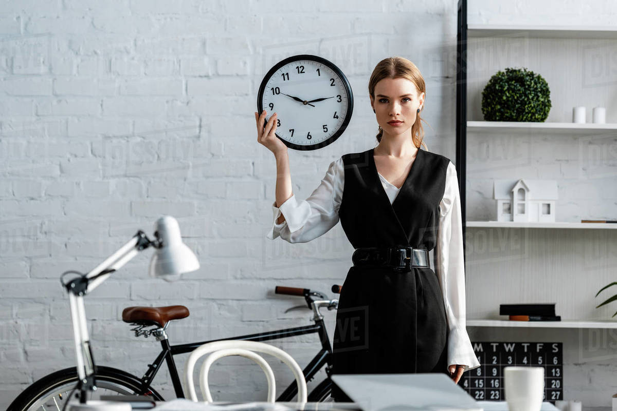 Serious businesswoman in formal wear holding clock at workplace - Stock ...