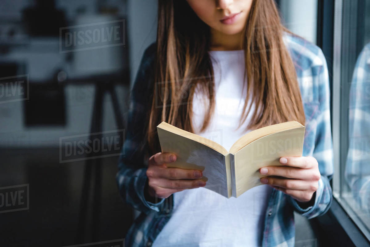 Cropped shot of young woman standing near window and reading book at ...
