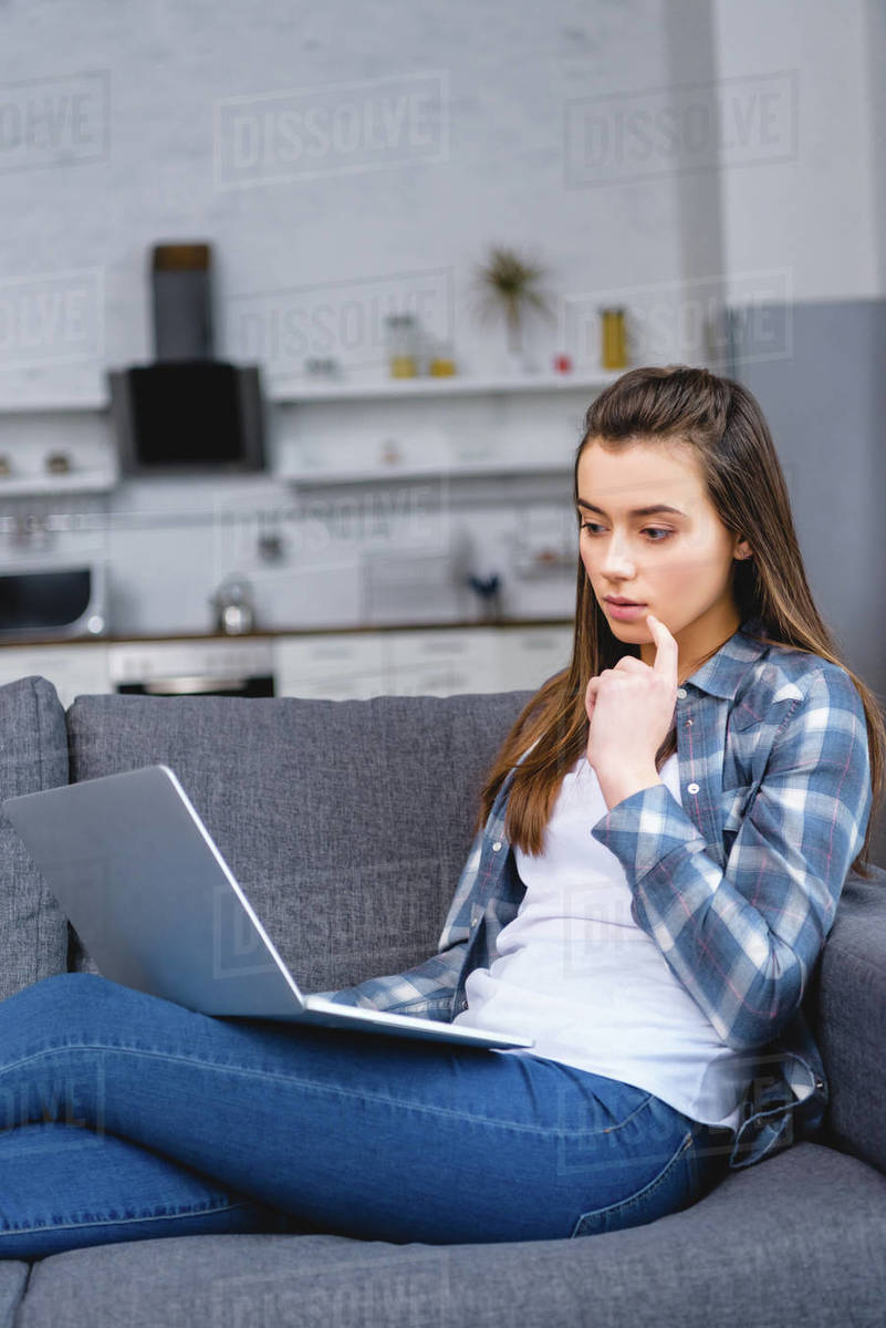 Thoughtful young woman sitting on couch and using laptop - Royalty-free ...