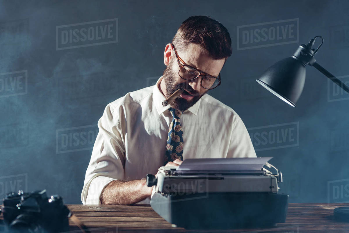 Bearded journalist sitting at table, typing on vintage typewriter and ...