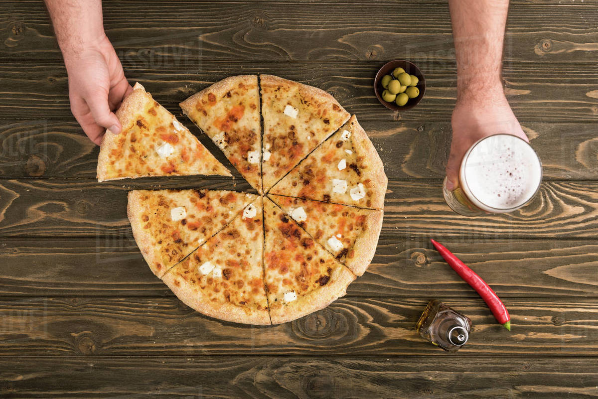 Cropped view of man eating cheese pizza on wooden table with beer and ...