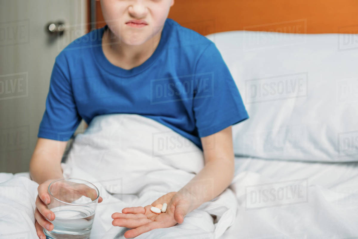 Unsatisfied boy holding glass of water and medicines in hospital bed ...