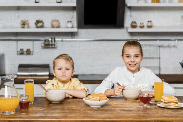 Pretty siblings looking at camera and sitting at table with cookies ...