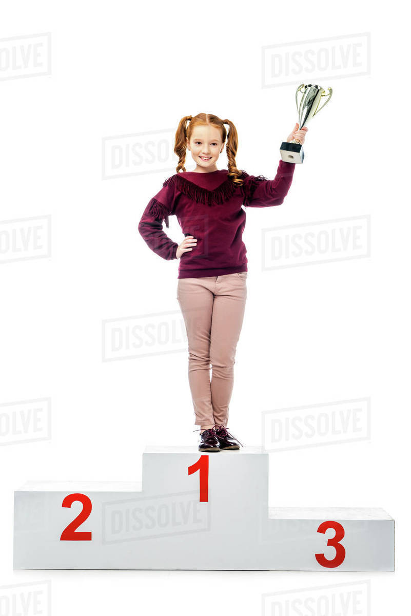 Cheerful schoolgirl standing on winner podium, smiling, holding trophy ...