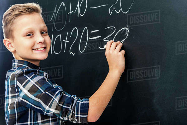Smiling boy writing math example on blackboard with chalk and looking ...