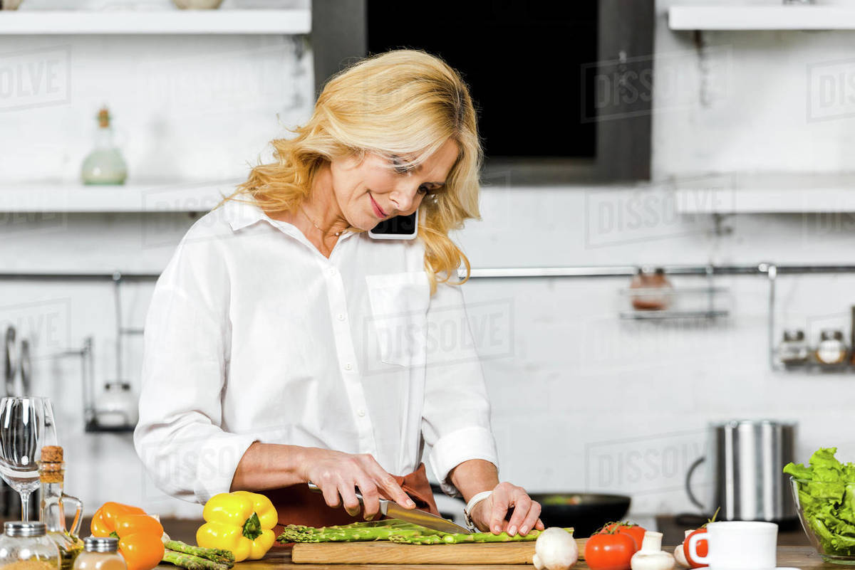 Attractive middle aged woman talking by smartphone while cooking in ...