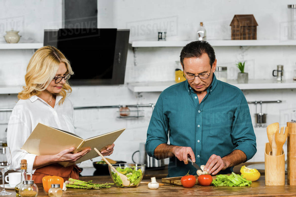 Middle aged wife and husband cooking together with recipe book in ...