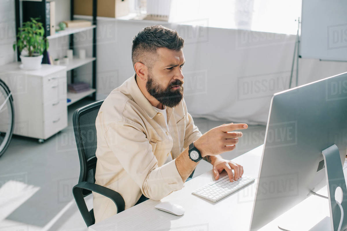 Concentrated businessman sitting at office desk and pointing at monitor ...