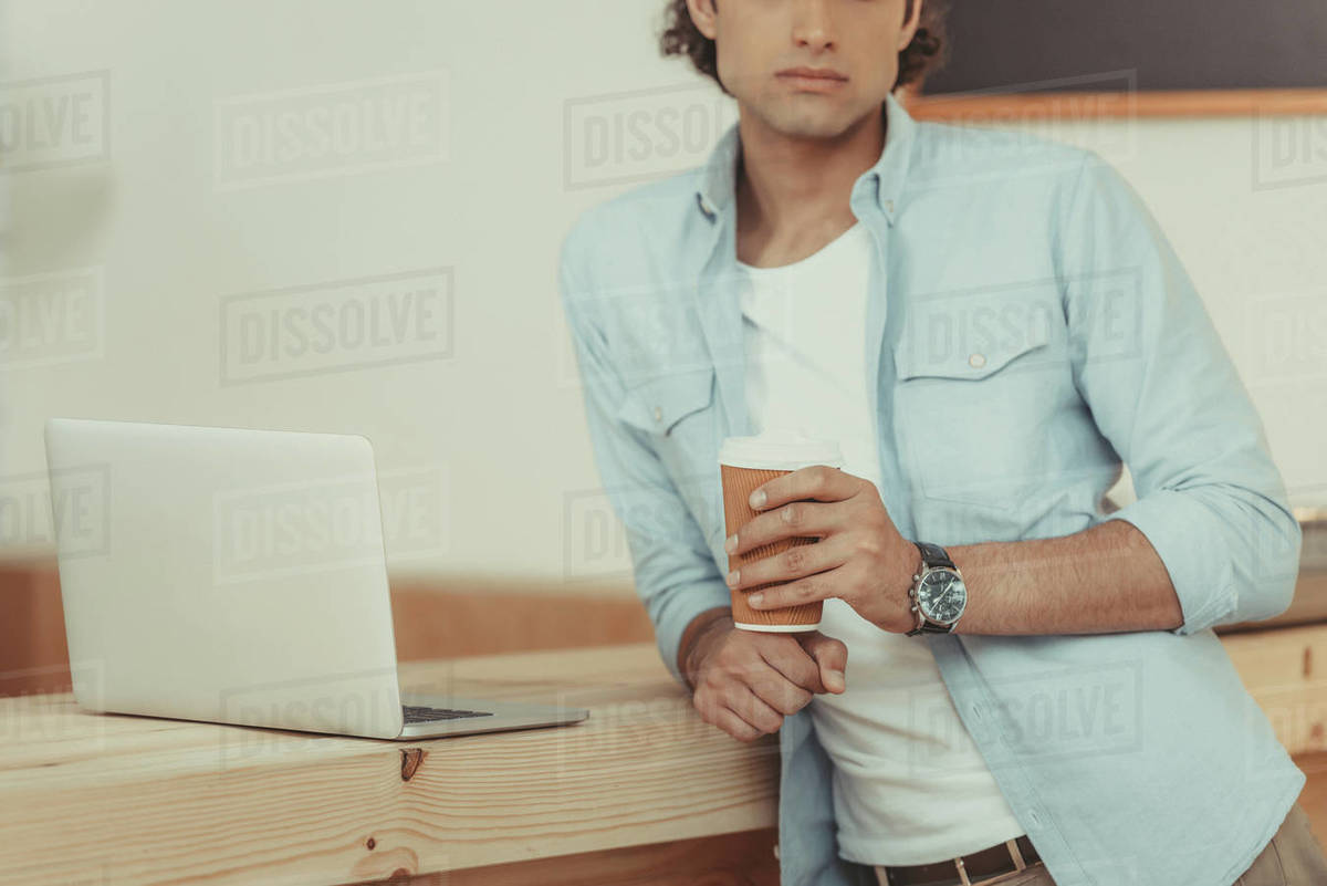Cropped shot of young man using laptop and drinking coffee in paper cup ...