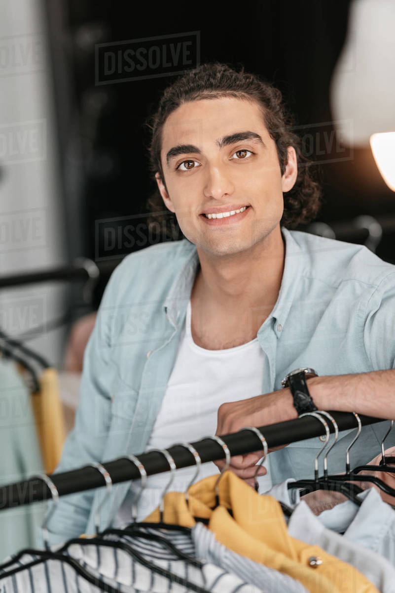 Handsome young boutique owner leaning at hangers with clothes and ...