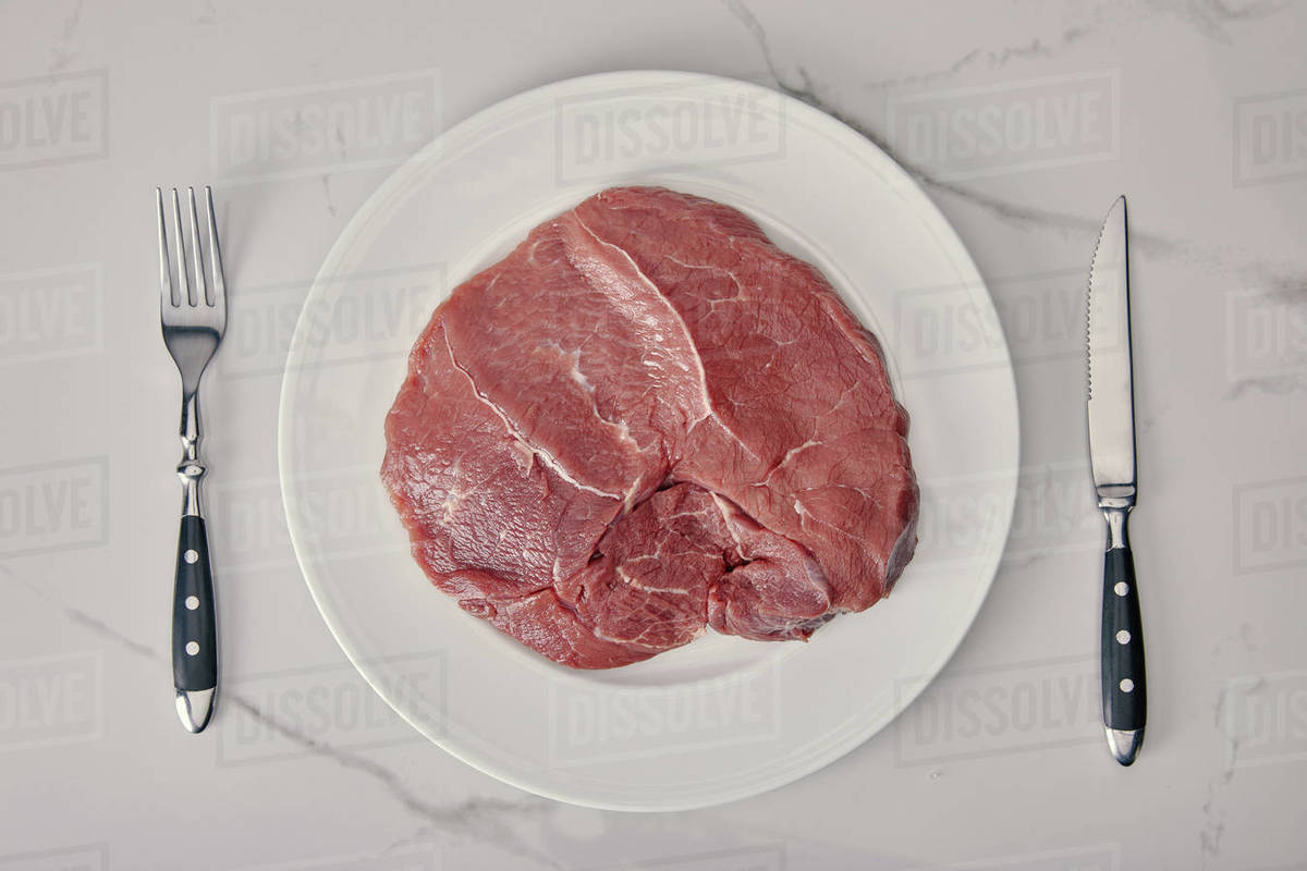 Top view of fresh raw meat on plate with kitchen cutlery on white ...