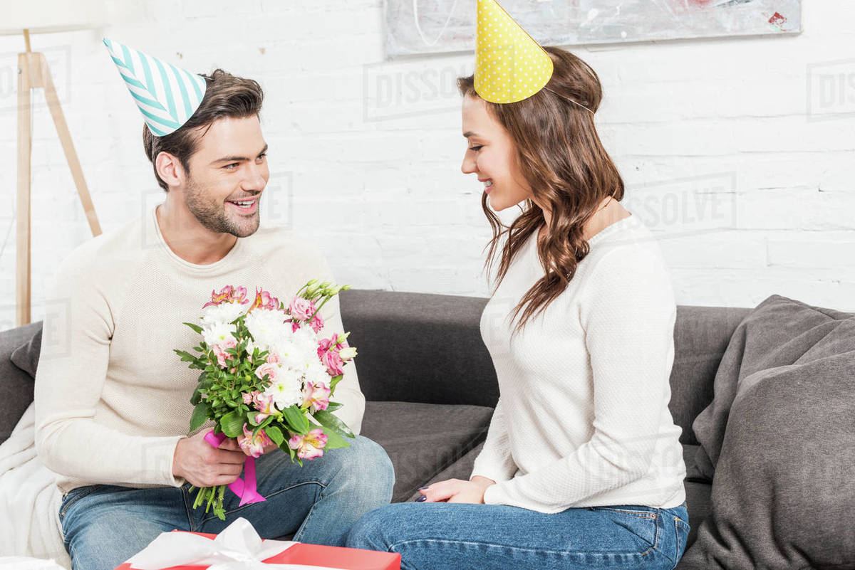 Handsome smiling man in party hat presenting birthday flowers to woman ...