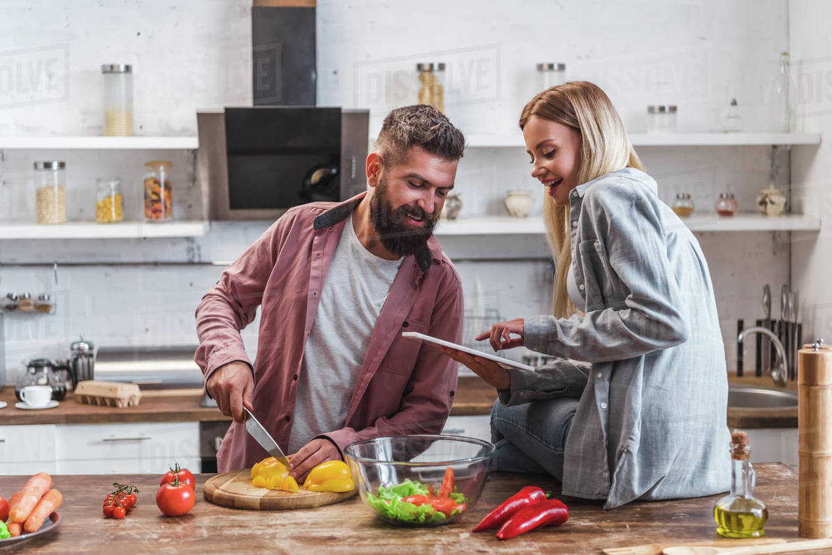 Husband making dinner and wife sitting on table with digital tablet ...
