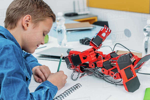 Schoolboy sitting at desk with robot model and writing in notebook ...