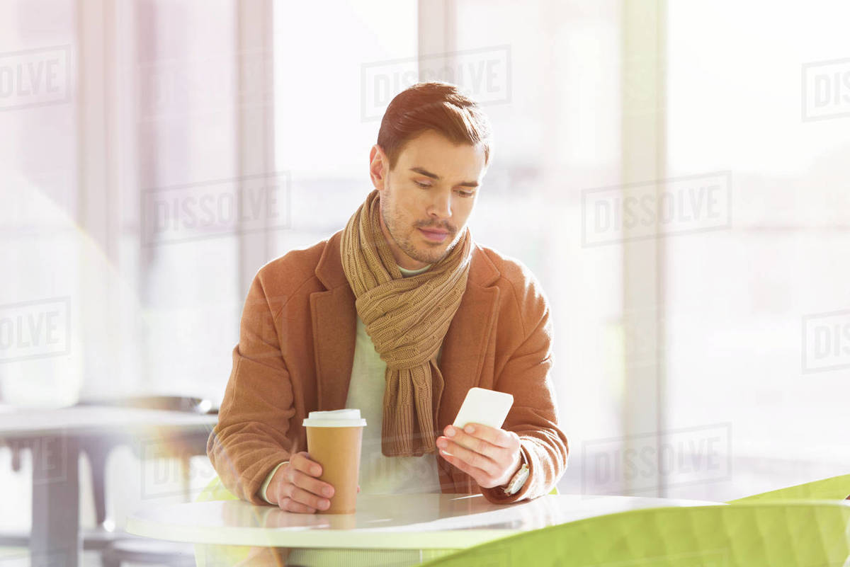 Handsome man sitting at table and holding smartphone and paper cup in ...
