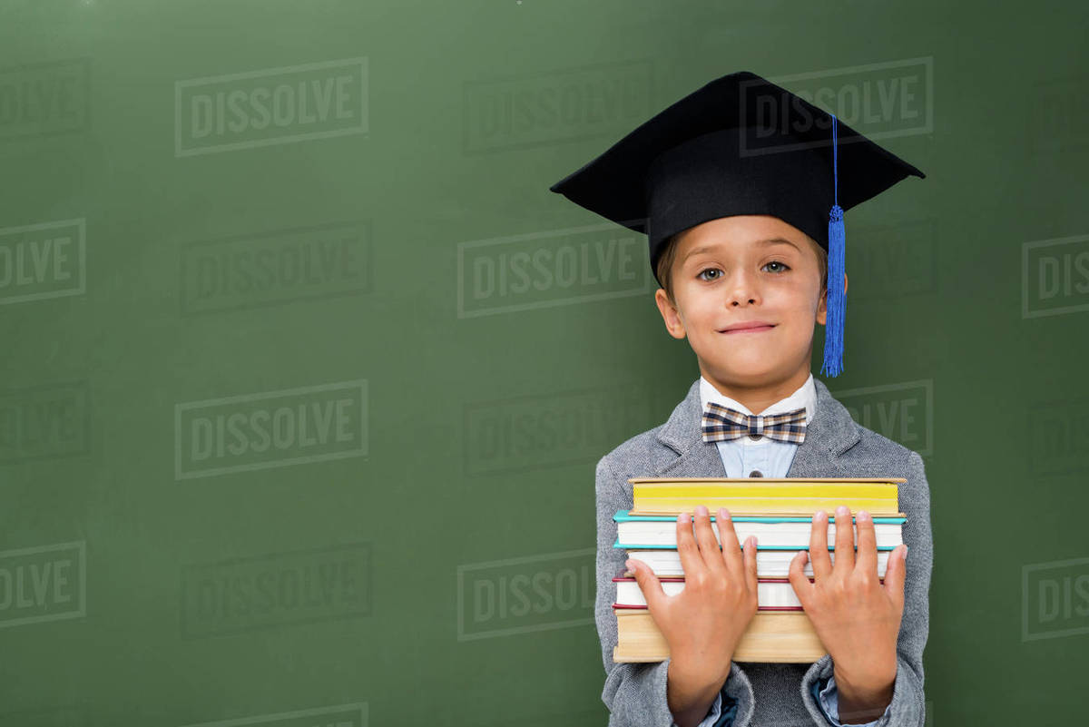 Schoolboy in graduation hat with stack of books next to chalkboard ...