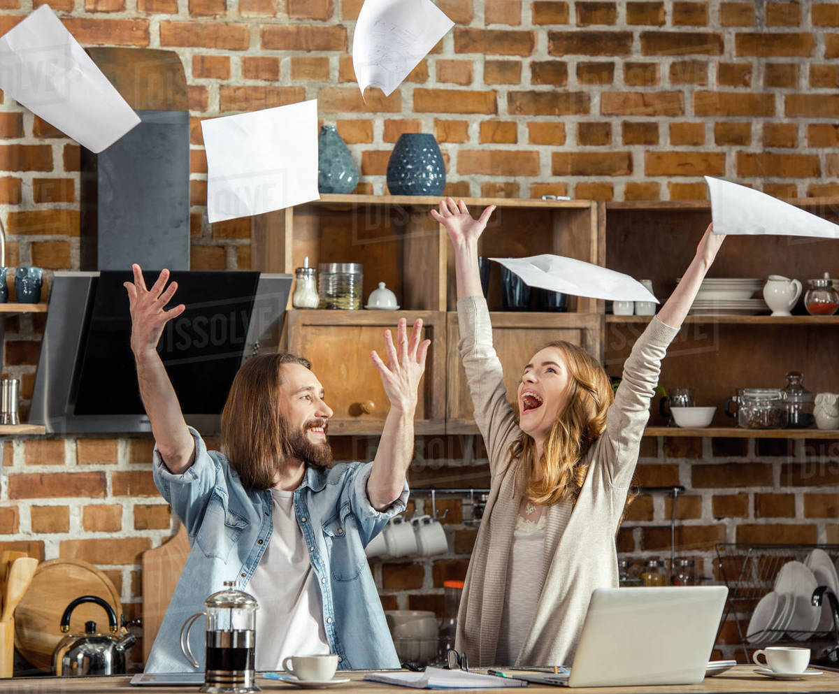 Excited young couple throwing documents while sitting and working with ...