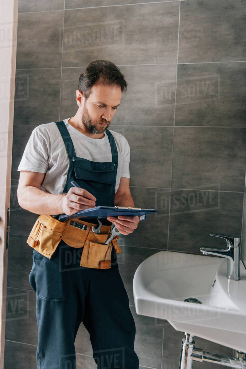 handsome male plumber with toolbelt writing in clipboard near broken ...