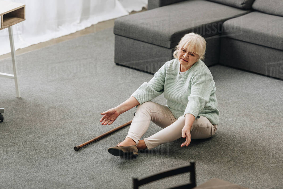 Helpless senior woman with blonde hair sitting on floor near sofa ...