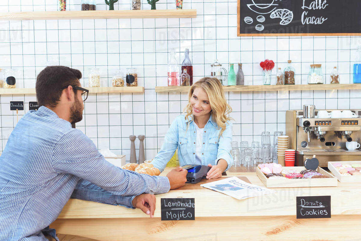 smiling young waitress with payment terminal looking at client in cafe ...