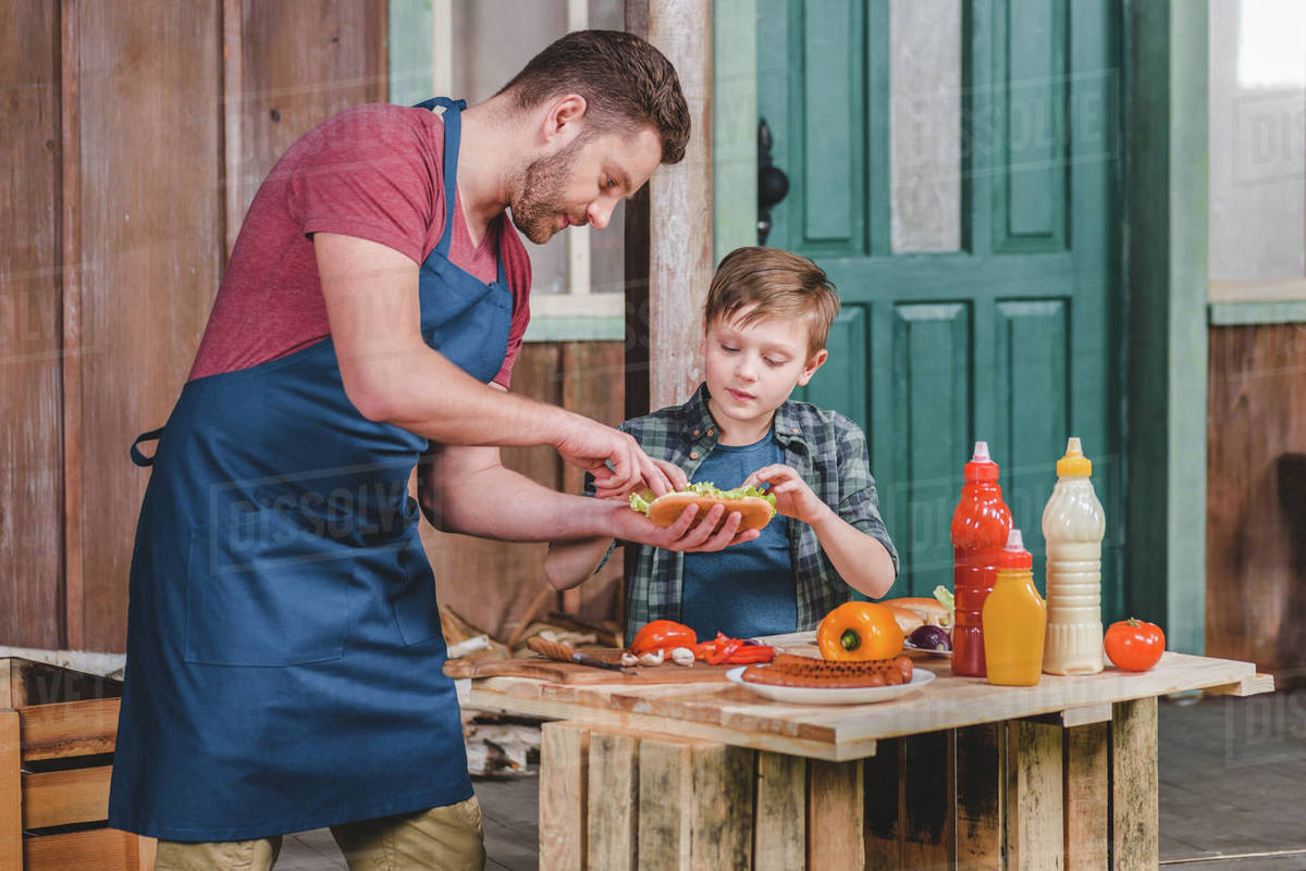 Smiling father and son preparing hot dog together in backyard, dad and ...