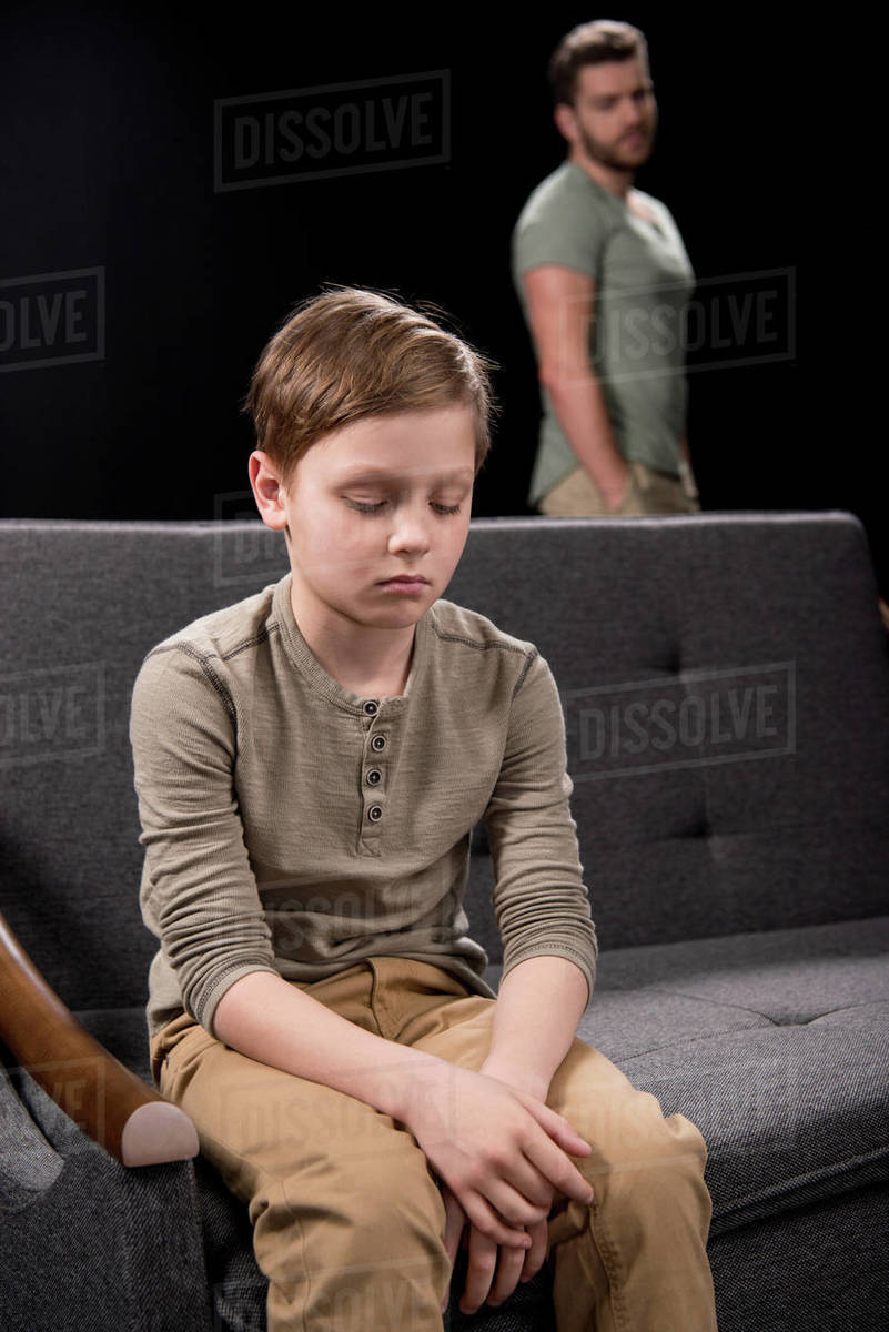 Upset little boy sitting on sofa and father standing behind, family ...
