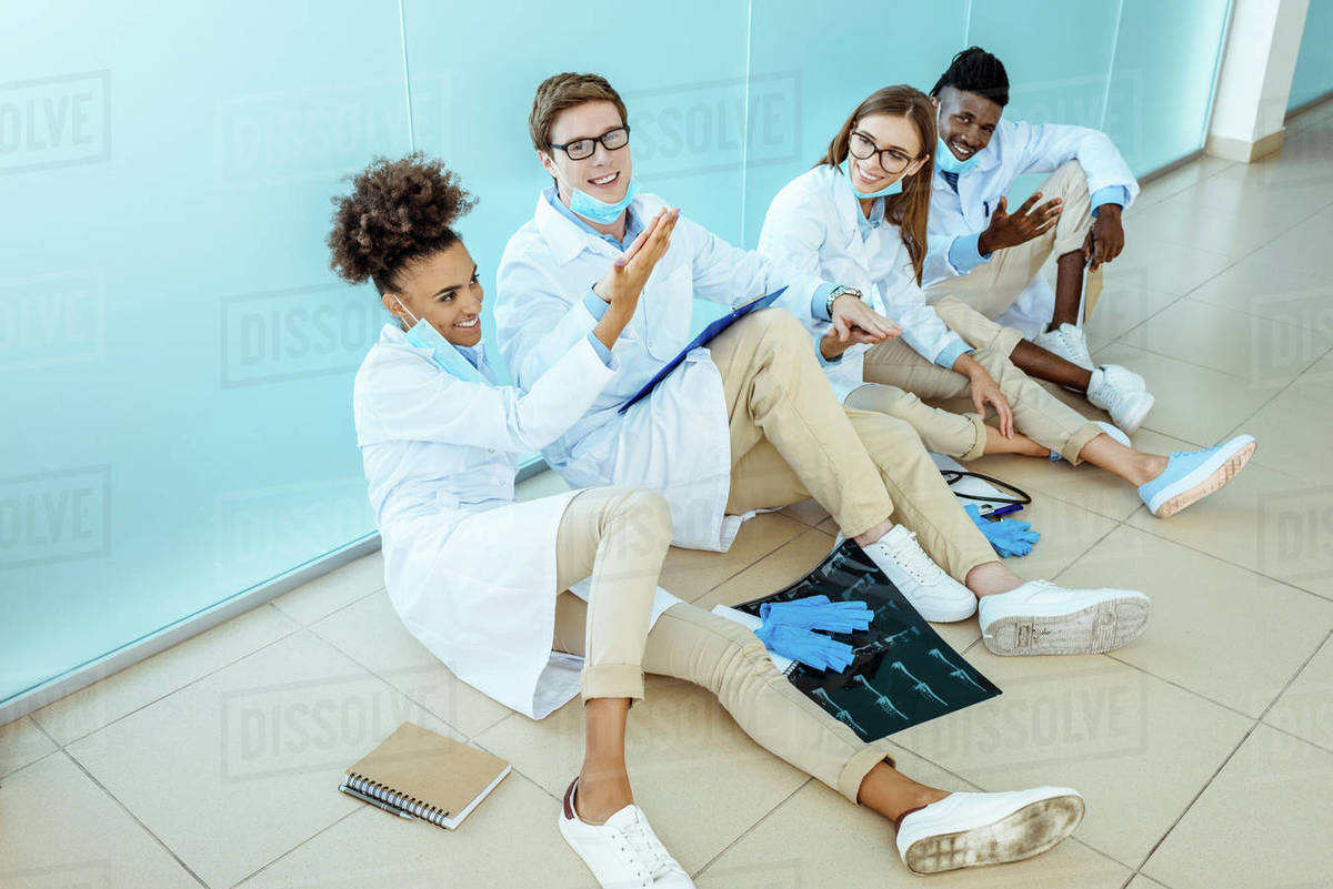 Four smiling young medical interns in white robes sitting on a floor in ...