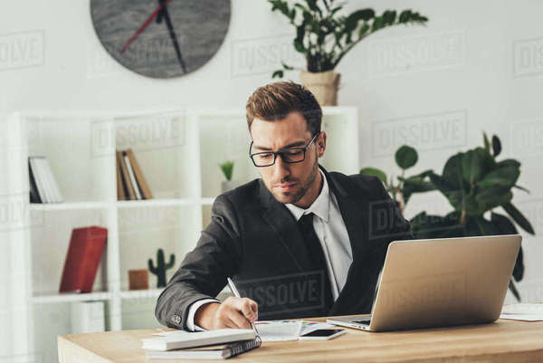 Young businessman writing in notebook while working with laptop - Stock ...