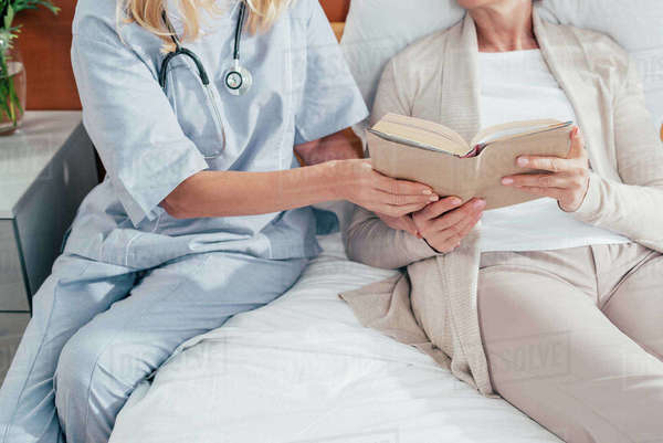 cropped shot of nurse and senior patient reading book together ...