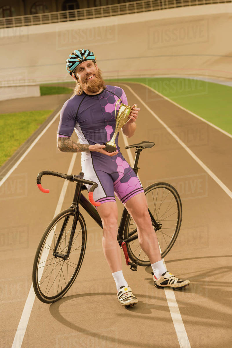 Smiling cyclist with champion cup looking at camera on cycle race track ...