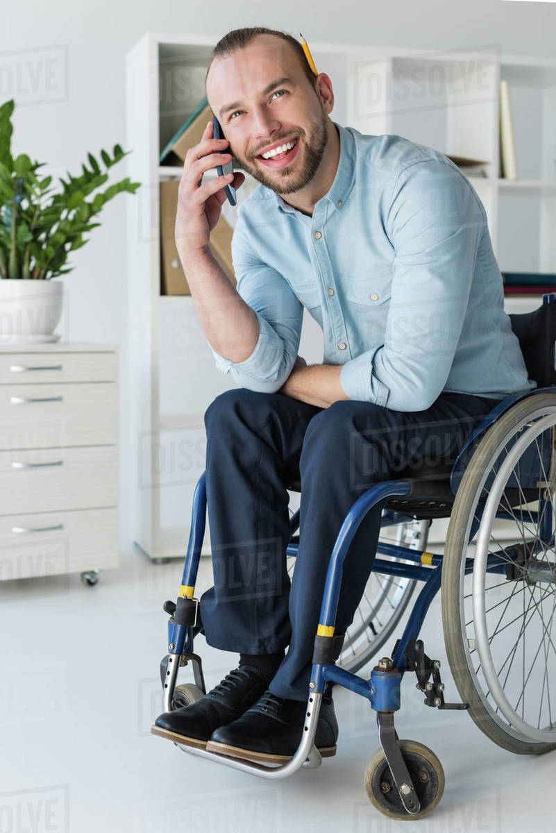 Smiling young man in wheelchair talking on smartphone in office ...