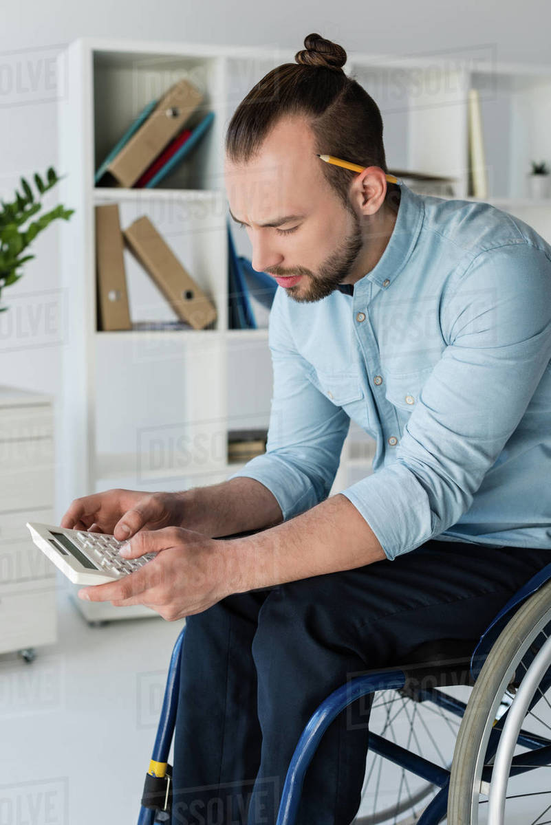 Smiling disabled businessman in wheelchair using calculator in office ...