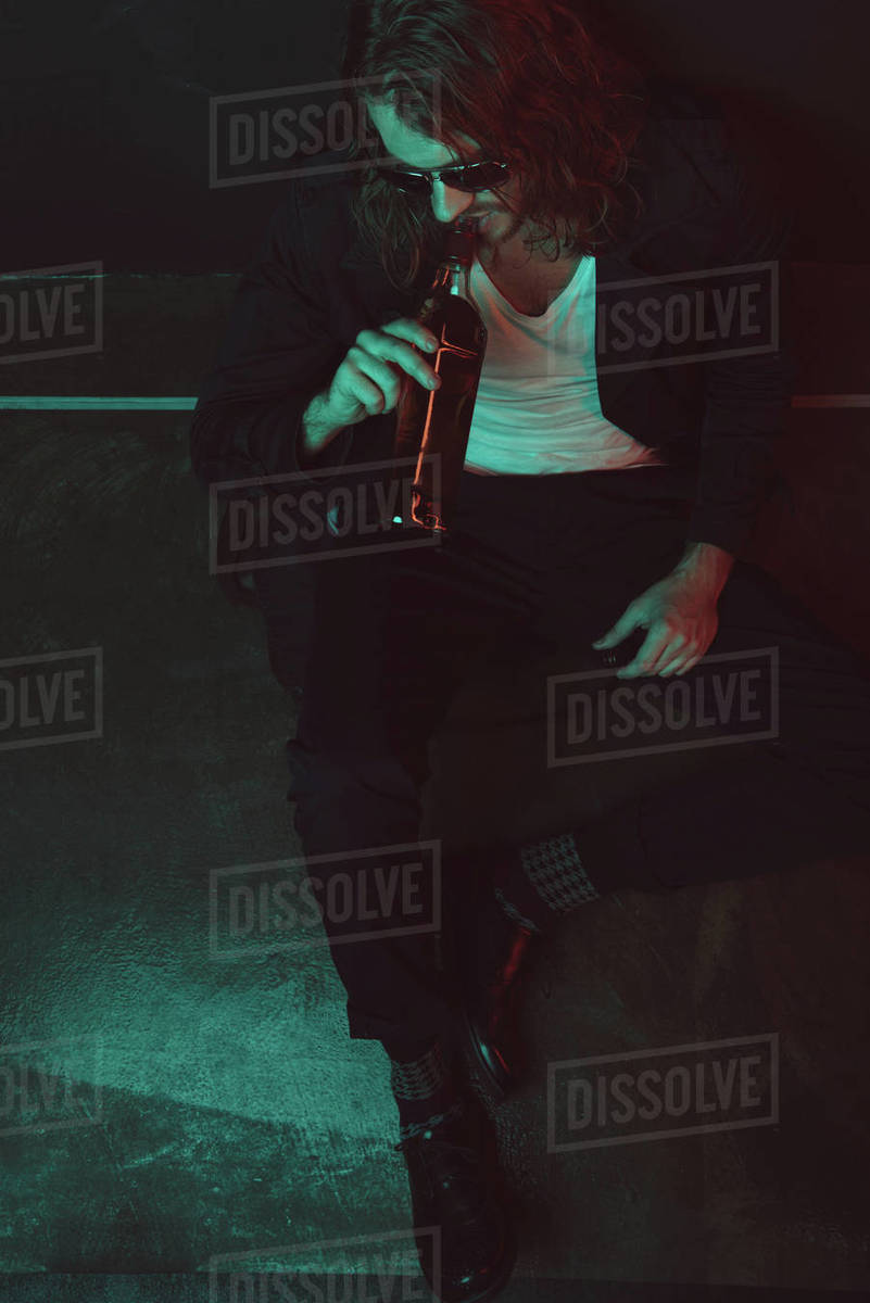 Young boozer in sunglasses drinking alcohol from bottle while sitting ...