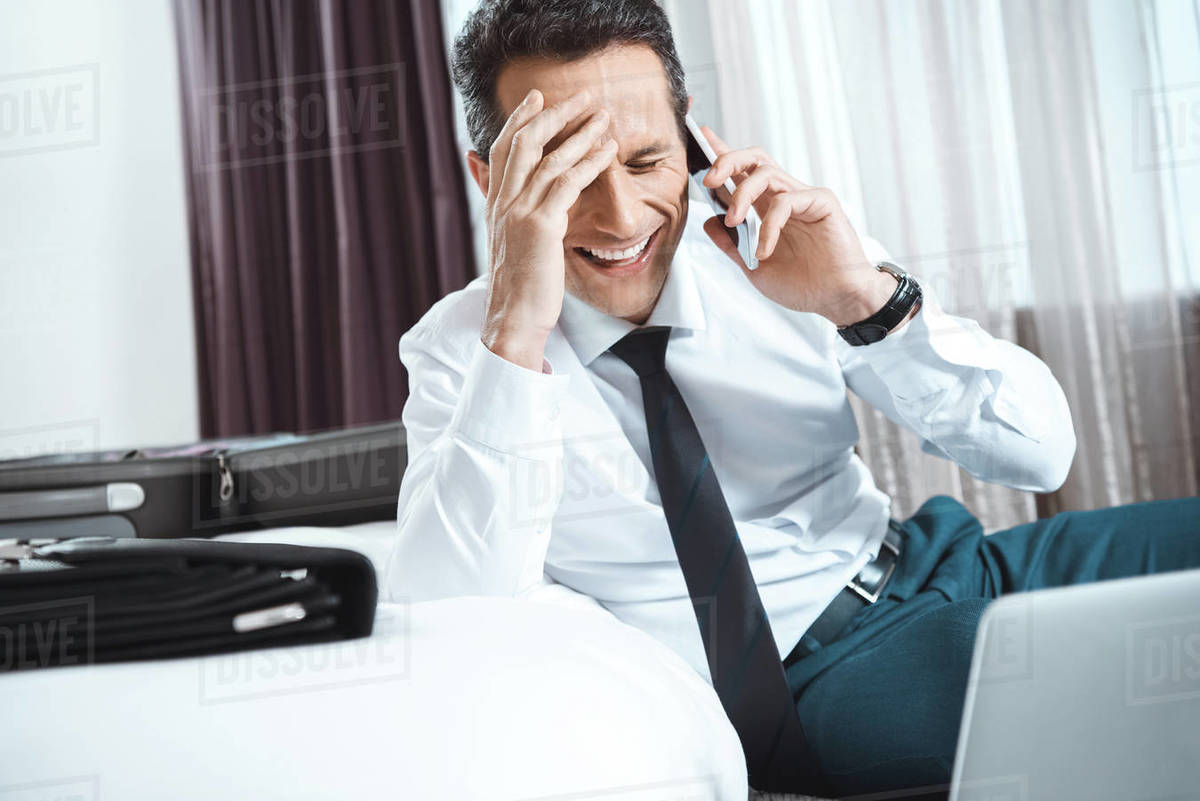 Laughing businessman in formal wear talking on phone while sitting near ...