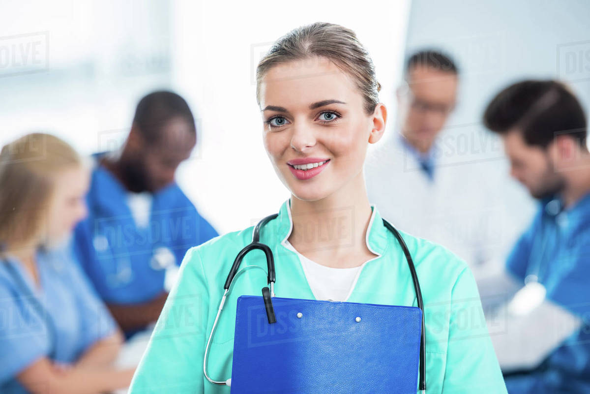 Smiling young nurse holding clipboard with colleagues sitting at table ...