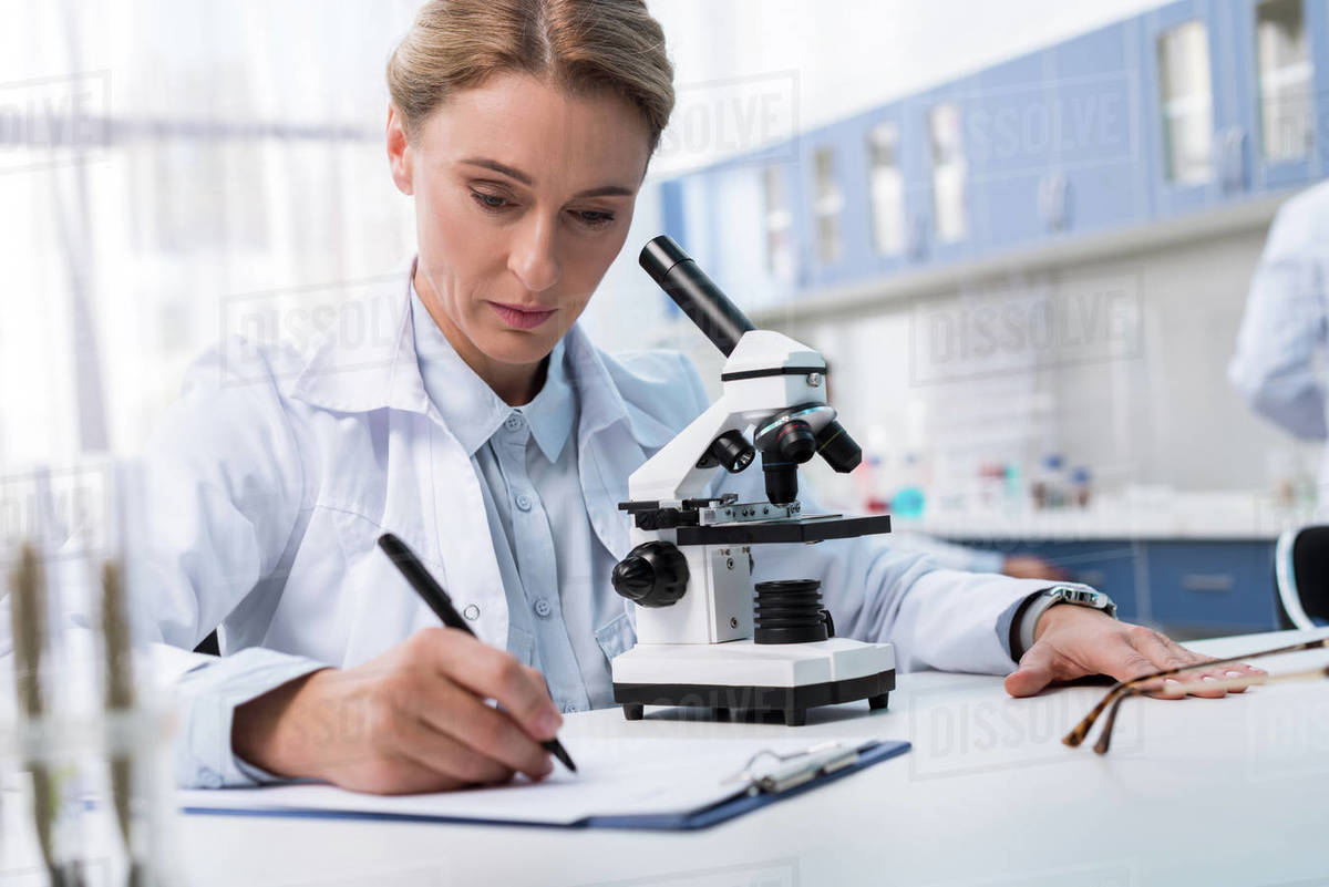 Professional lab technician taking notes while doing microscope sample ...
