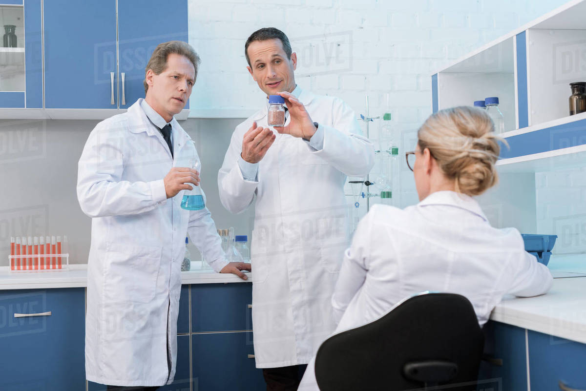 Two professional scientists showing their colleague jars with chemicals ...