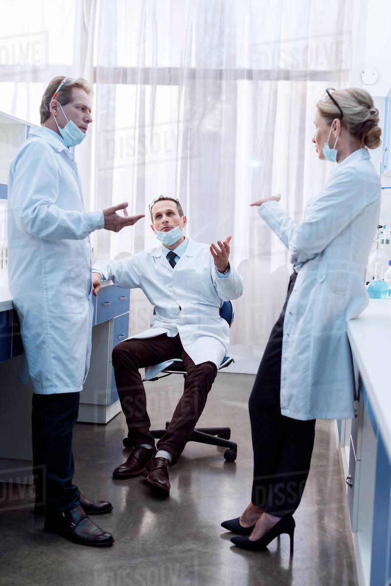 Three medical workers in lab coats and sterile masks discussing work in ...
