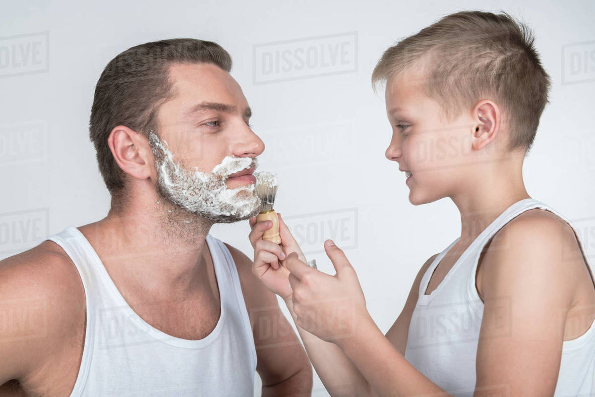 Cute little boy shaving father with shaving foam isolated on grey ...