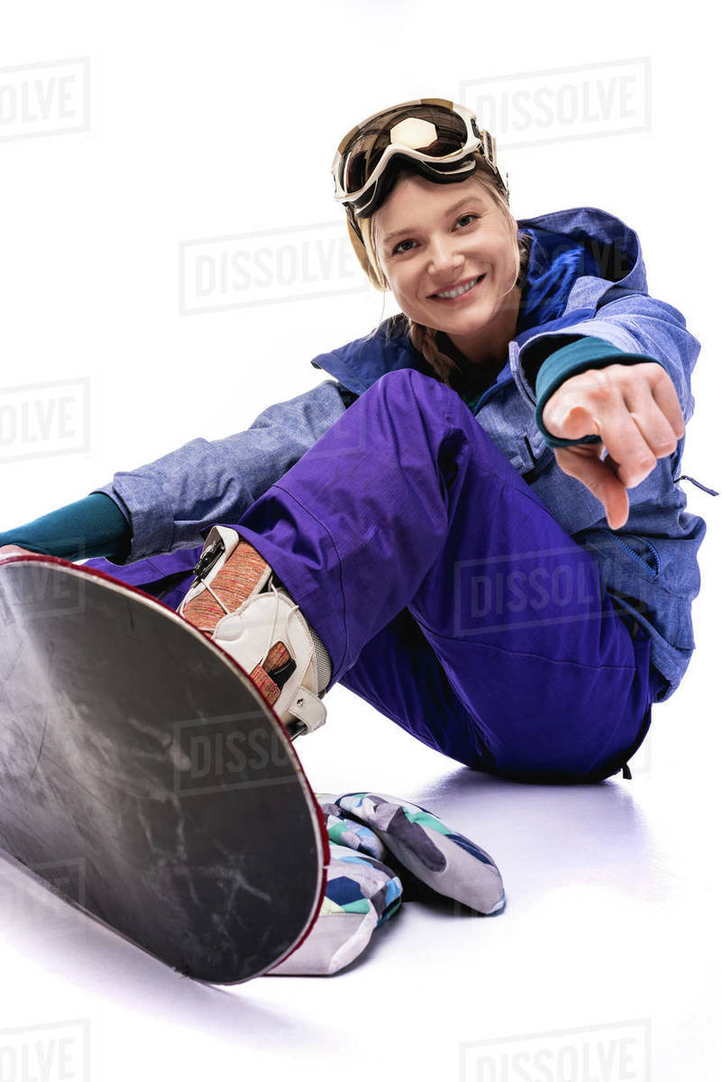 Selective focus of smiling snowboarder pointing at camera isolated on ...