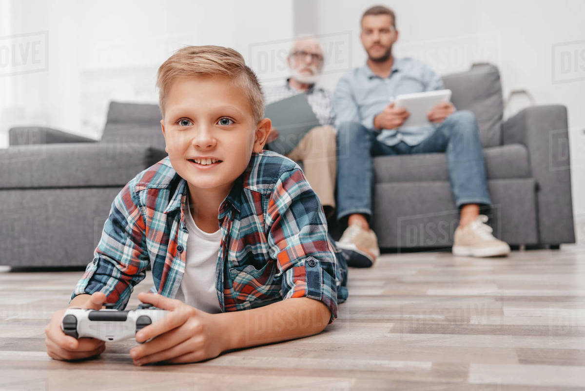 Little boy lying with gamepad on floor in living room, playing videogames while his father and ...