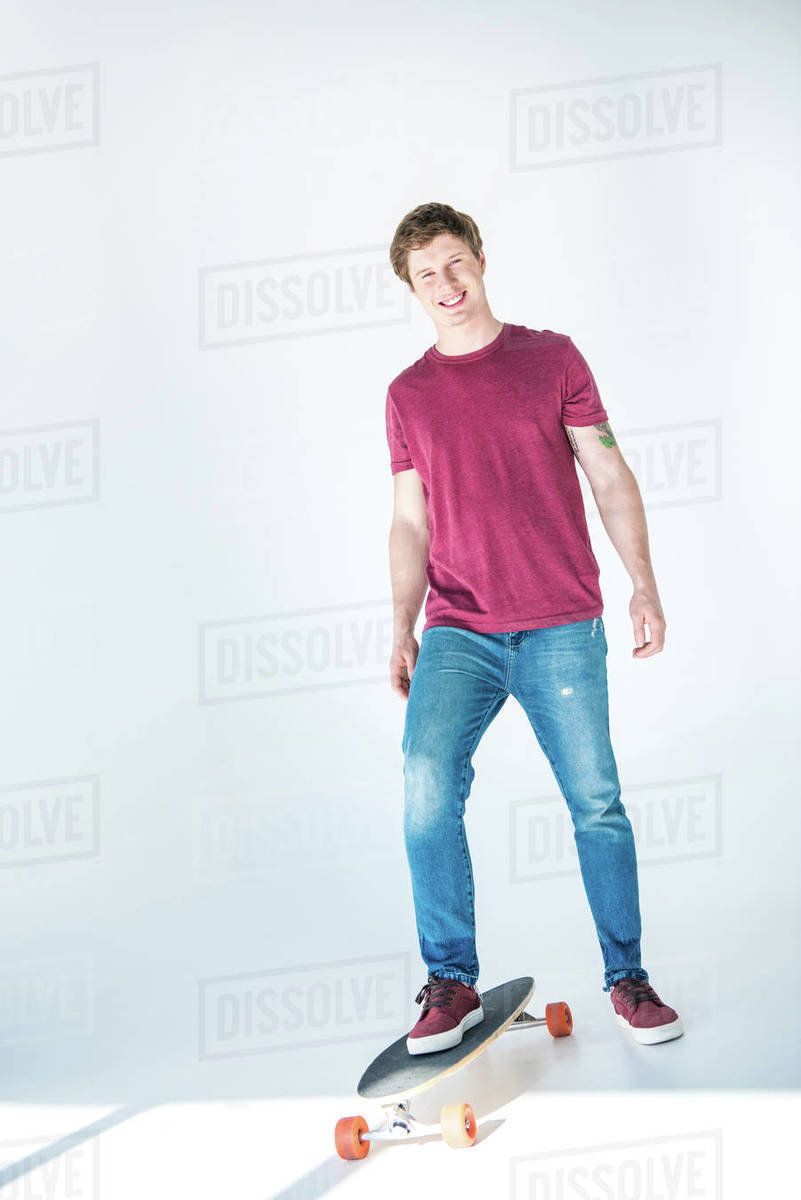 Handsome young man standing on skateboard and looking at camera on grey ...