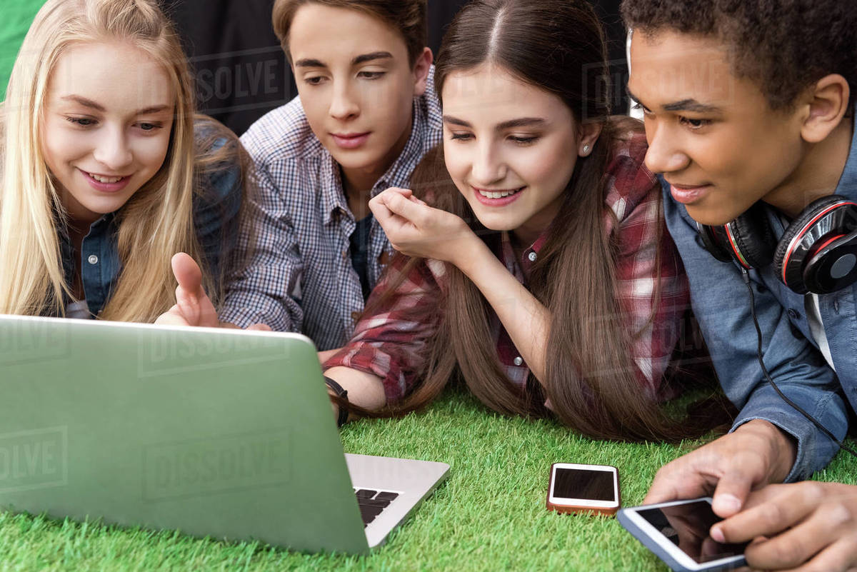 Portrait of multicultural smiling teenagers using laptop together while ...